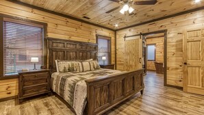 Quarto de madeira com cama, mesas de cabeceira, ventilador de teto e porta de madeira.