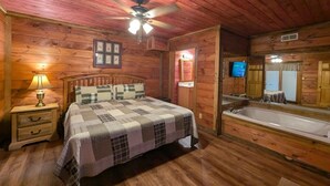Quarto de cabana de madeira com cama, mesa de cabeceira, abajur, ventoinha no teto, banheiro com banheira e televisão.