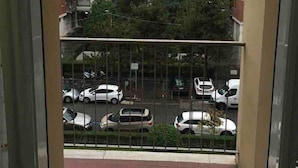 Vista de uma varanda sobre um parque de estacionamento com vários carros, uma fileira de árvores e um prédio residencial de vários andares ao fundo.