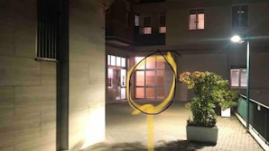 Vista noturna da fachada de um edifício com um letreiro circular, plantas em vasos e área pavimentada com tijolos.
