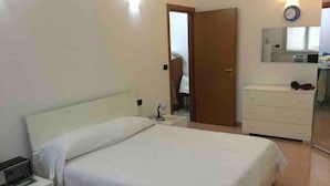 Quarto com cama, mesa de cabeceira, cômoda, espelho e porta que dá para outro espaço.