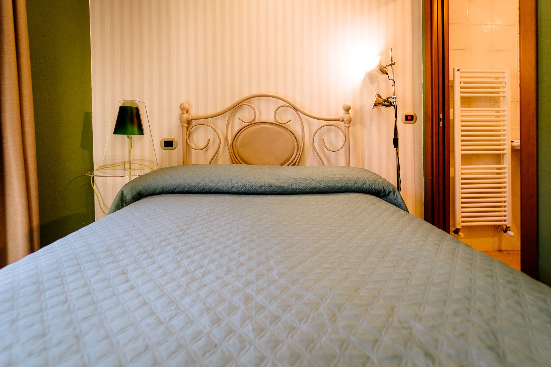 Cama bem arrumada com cabeceira acolchoada, luminária de cabeceira e termostato na parede.
