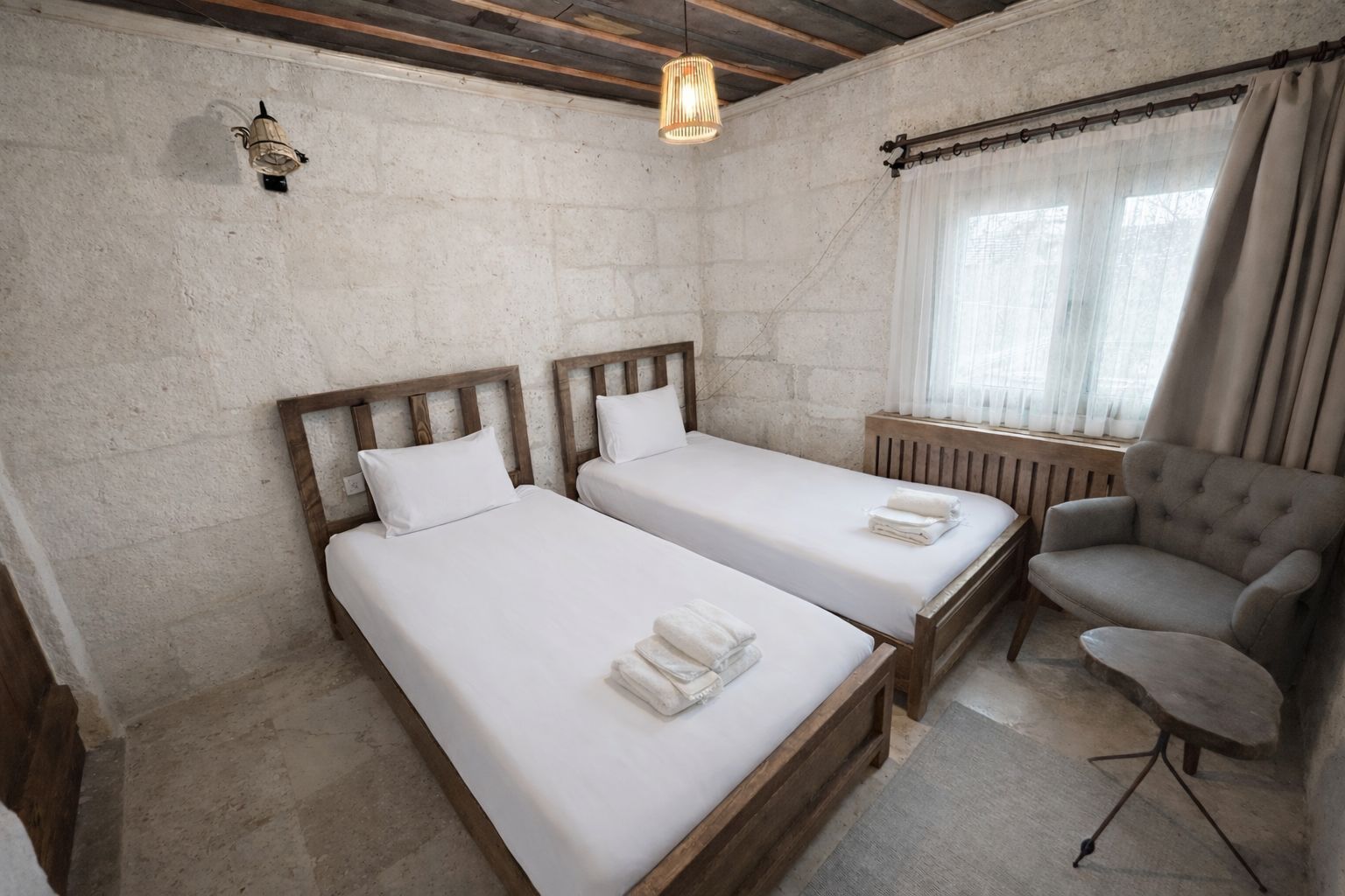 Quarto com duas camas de solteiro, uma cadeira de madeira, um radiador e uma janela com cortinas.