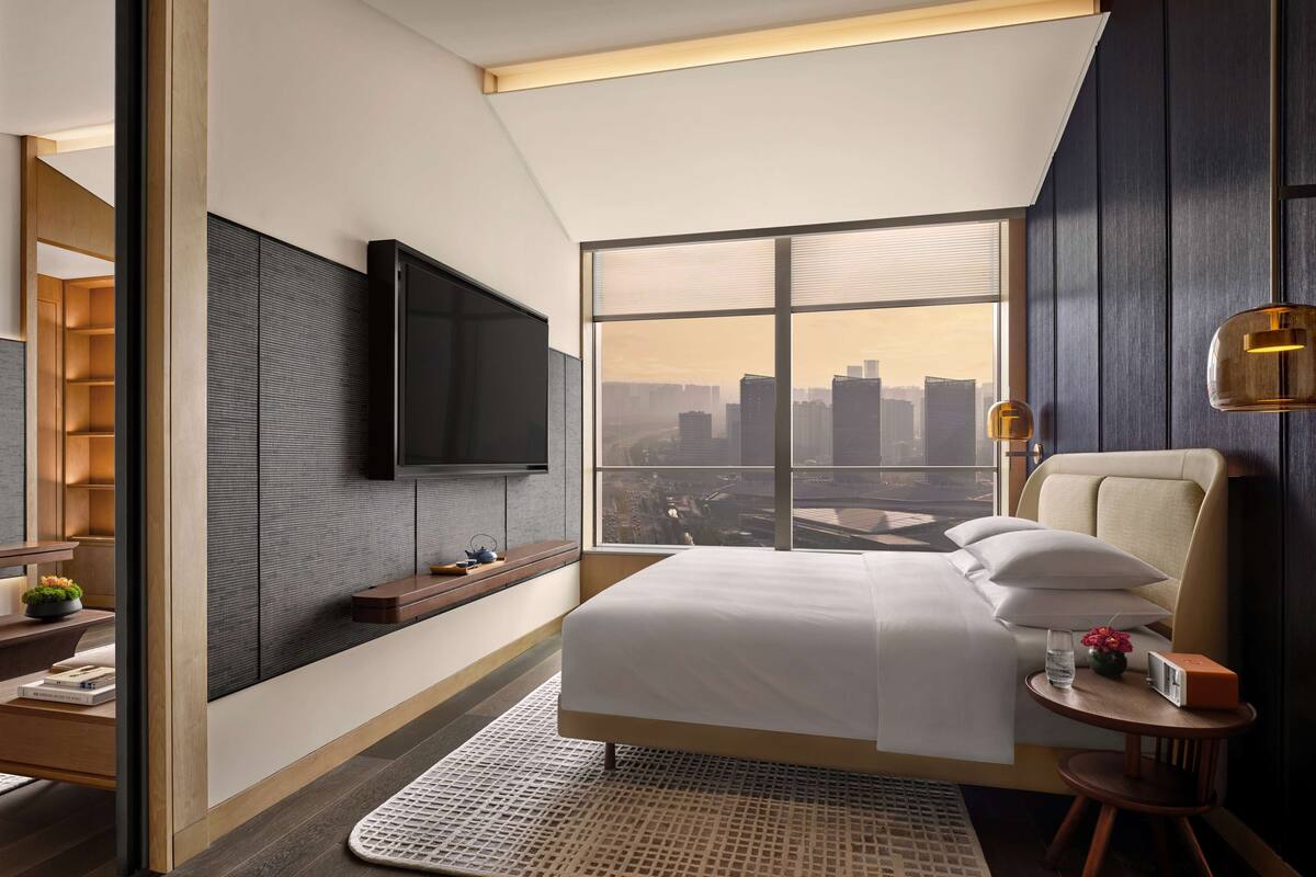 Quarto de hotel moderno com uma cama grande, uma televisão presa à parede, vista para a paisagem urbana e uma pequena mesa com abajur.