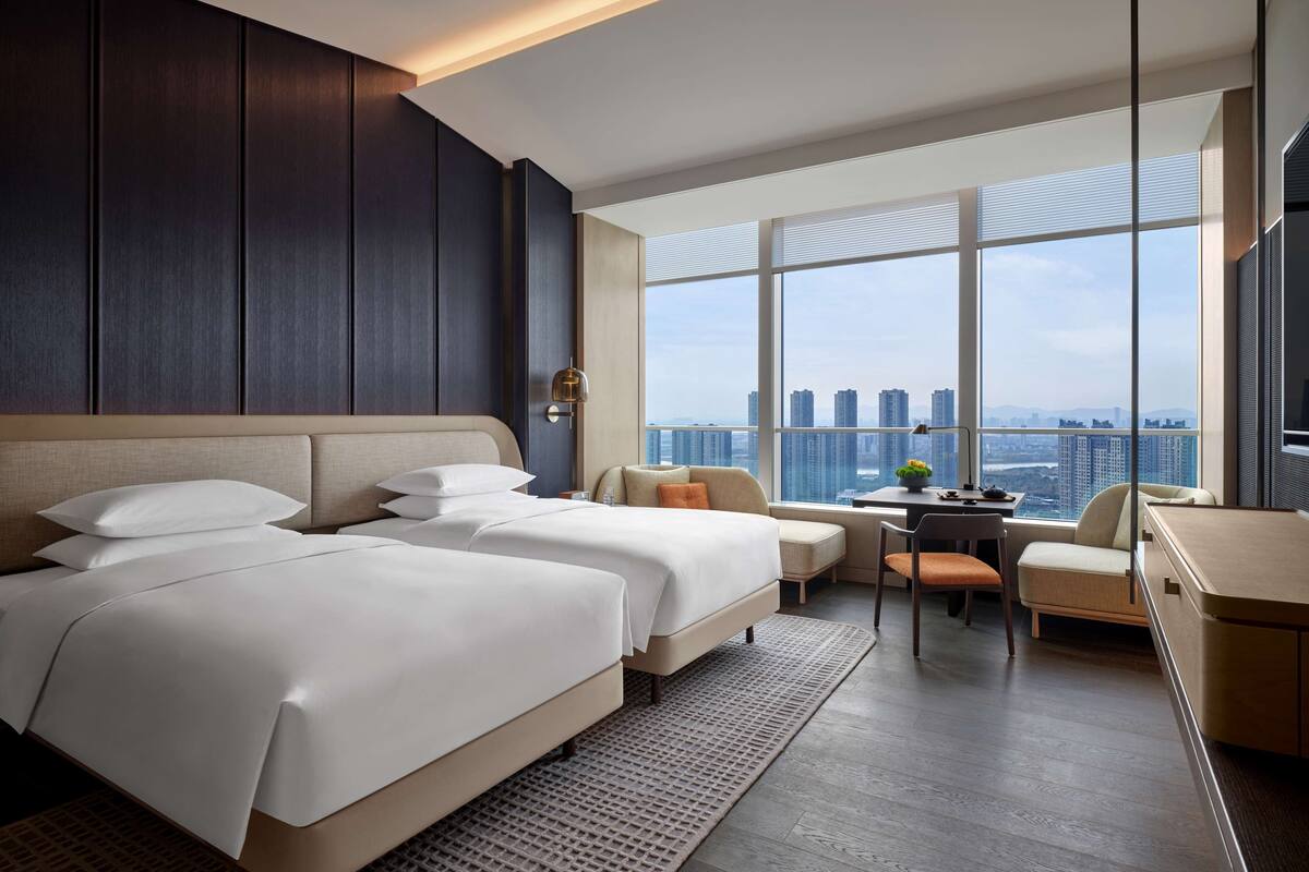 Quarto de hotel moderno com duas camas grandes, uma mesinha, uma cadeira e vista para o horizonte da cidade.
