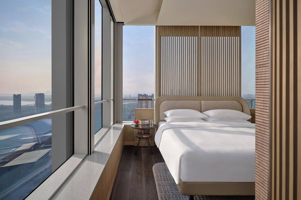Quarto de hotel moderno com uma cama grande, uma mesinha com um vaso e vista da cidade através de janelas do chão ao teto.