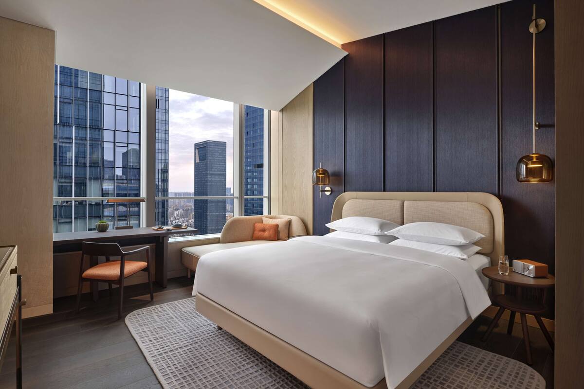 Quarto de hotel moderno com uma cama grande, uma escrivaninha e vista para a cidade através de janelas amplas.