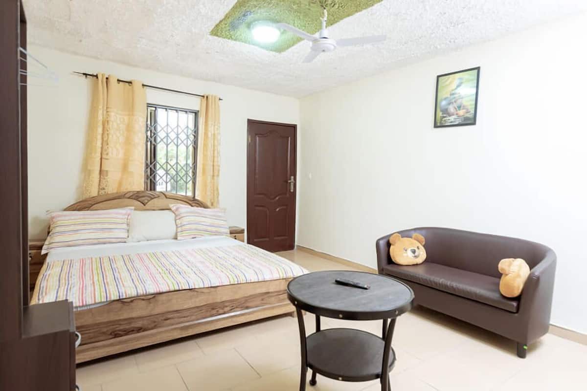 Quarto com cama, uma mesinha redonda, um sofá com um urso de pelúcia, janela com cortinas e uma porta.