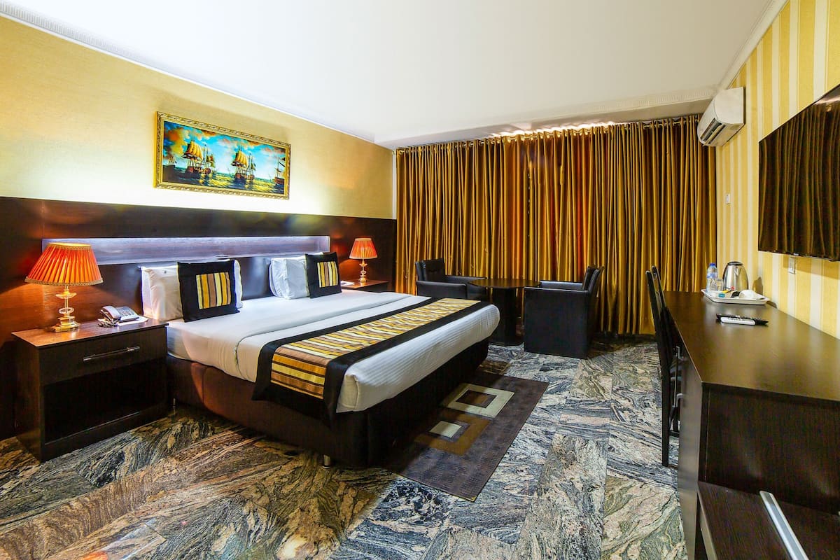 Quarto de hotel com uma cama grande, duas mesas de cabeceira com luminárias, uma escrivaninha com cadeira, televisão e uma grande janela com cortinas.