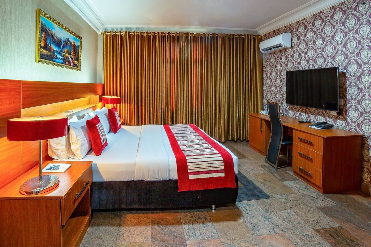 Quarto de hotel com cama, escrivaninha, televisão e um quadro na parede.