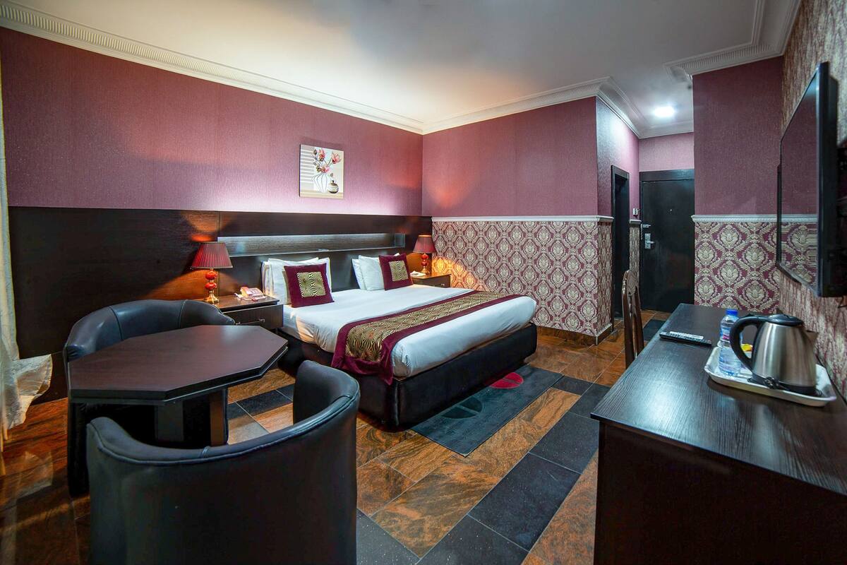 Quarto de hotel com cama, escrivaninha, cadeira, televisão e porta do banheiro.