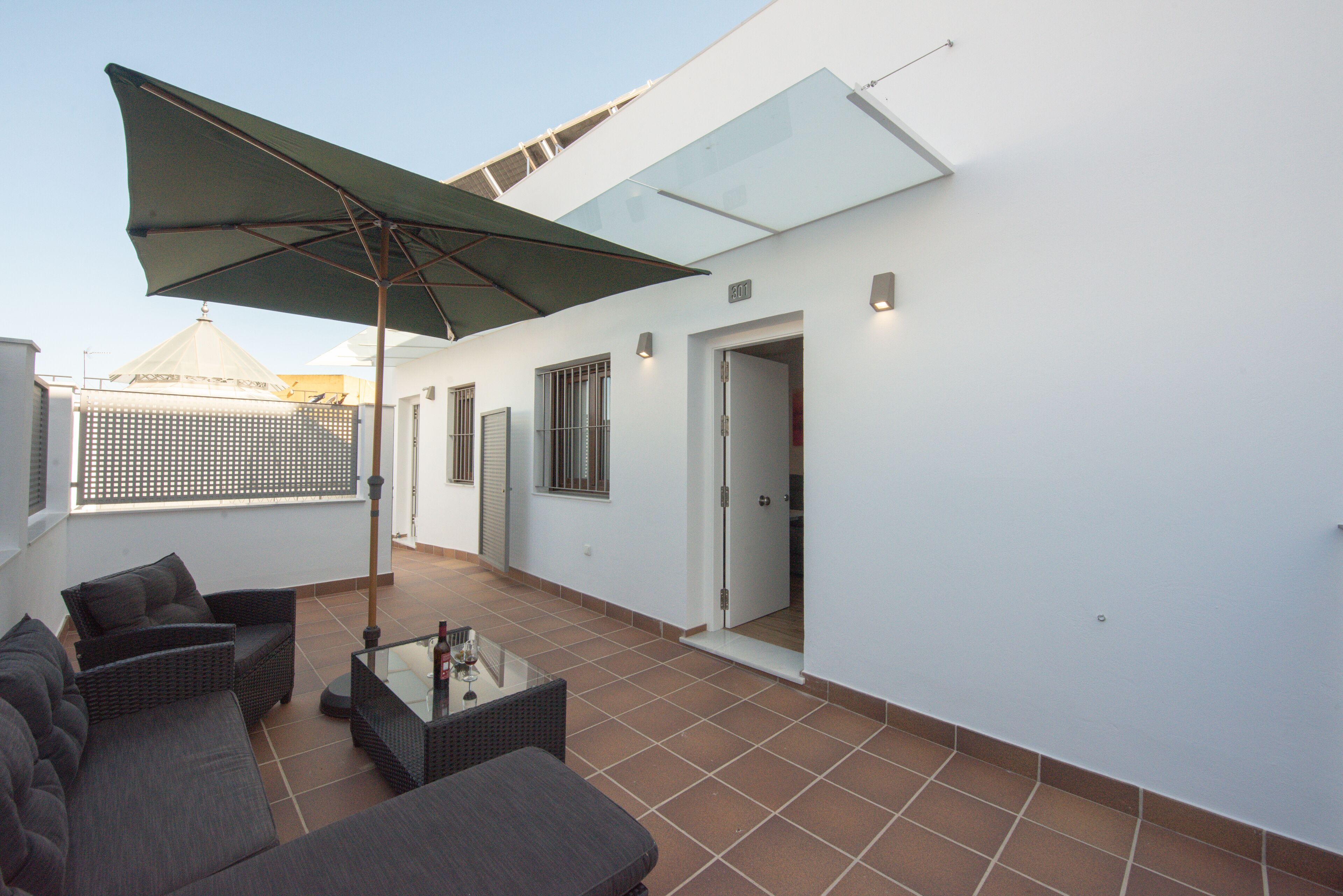Um terraço no telhado com um grande guarda-sol, móveis de vime e uma mesa de vidro.