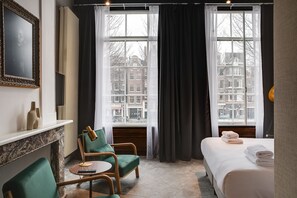 Quarto de hotel com cama, duas poltronas verdes, uma mesinha, lareira e grandes janelas com cortinas.