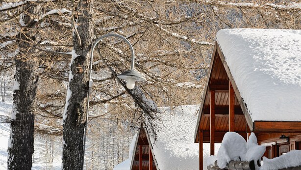 Uma cabana de madeira coberta por neve, com chaminé visível, rodeada por árvores e um caminho também coberto de neve.