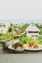 Refeição com iogurte, granola e um waffle numa varanda com vista para edifícios e vegetação.