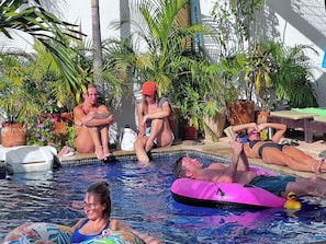 Área de piscina com várias pessoas, algumas na borda e outras boiando.