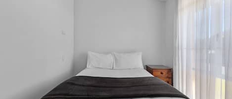 Uma cama bem arrumada com lençóis brancos e um edredom escuro, uma mesa de cabeceira de madeira e cortinas leves em uma janela.