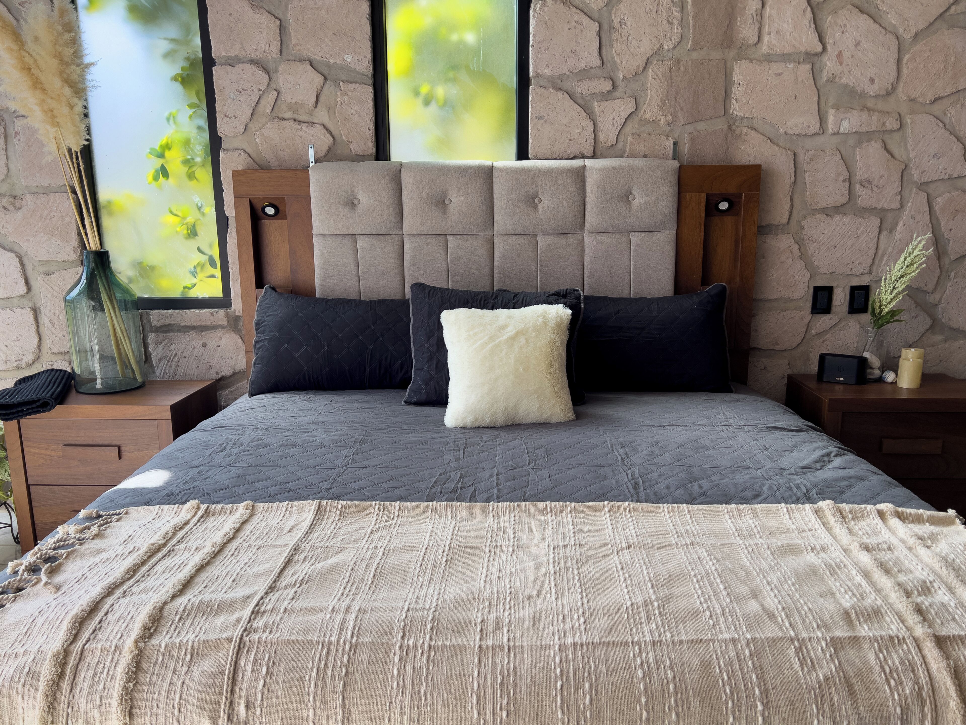 Uma cama com cabeceira estofada, duas almofadas pretas, uma almofada branca e fofa e um cobertor texturizado.