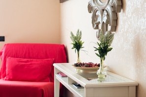 Uma sala de estar com um sofá vermelho, uma mesinha lateral branca com um recipiente de petiscos e dois vasos com plantas.