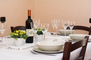 Mesa de jantar posta com pratos brancos, copos de vinho transparentes e uma garrafa de vinho. Há pequenas plantas em vasos e um jogo americano de renda.