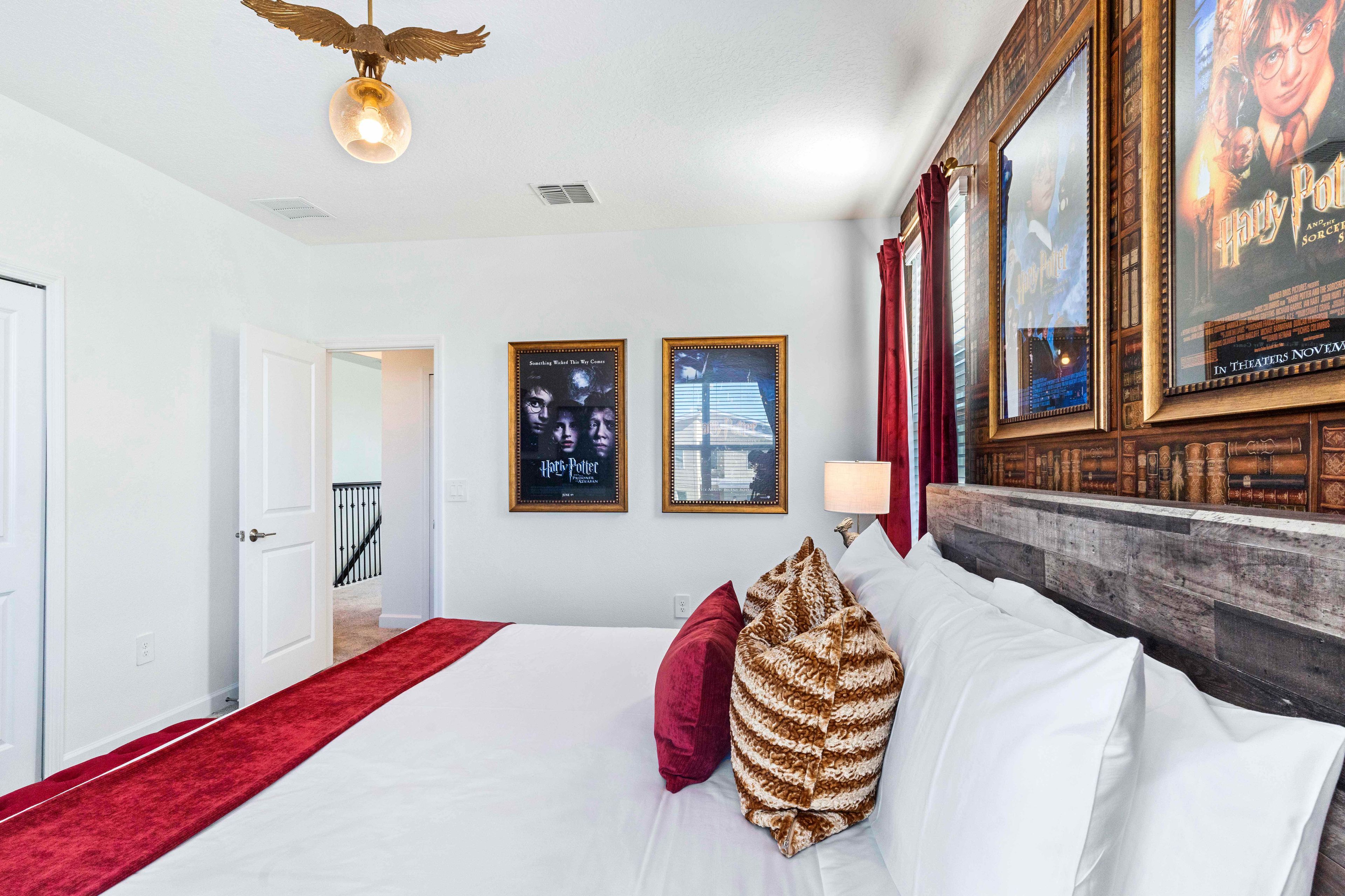 Quarto de hotel com uma cama grande, um tapete vermelho, cartazes emoldurados na parede e um lustre no teto.