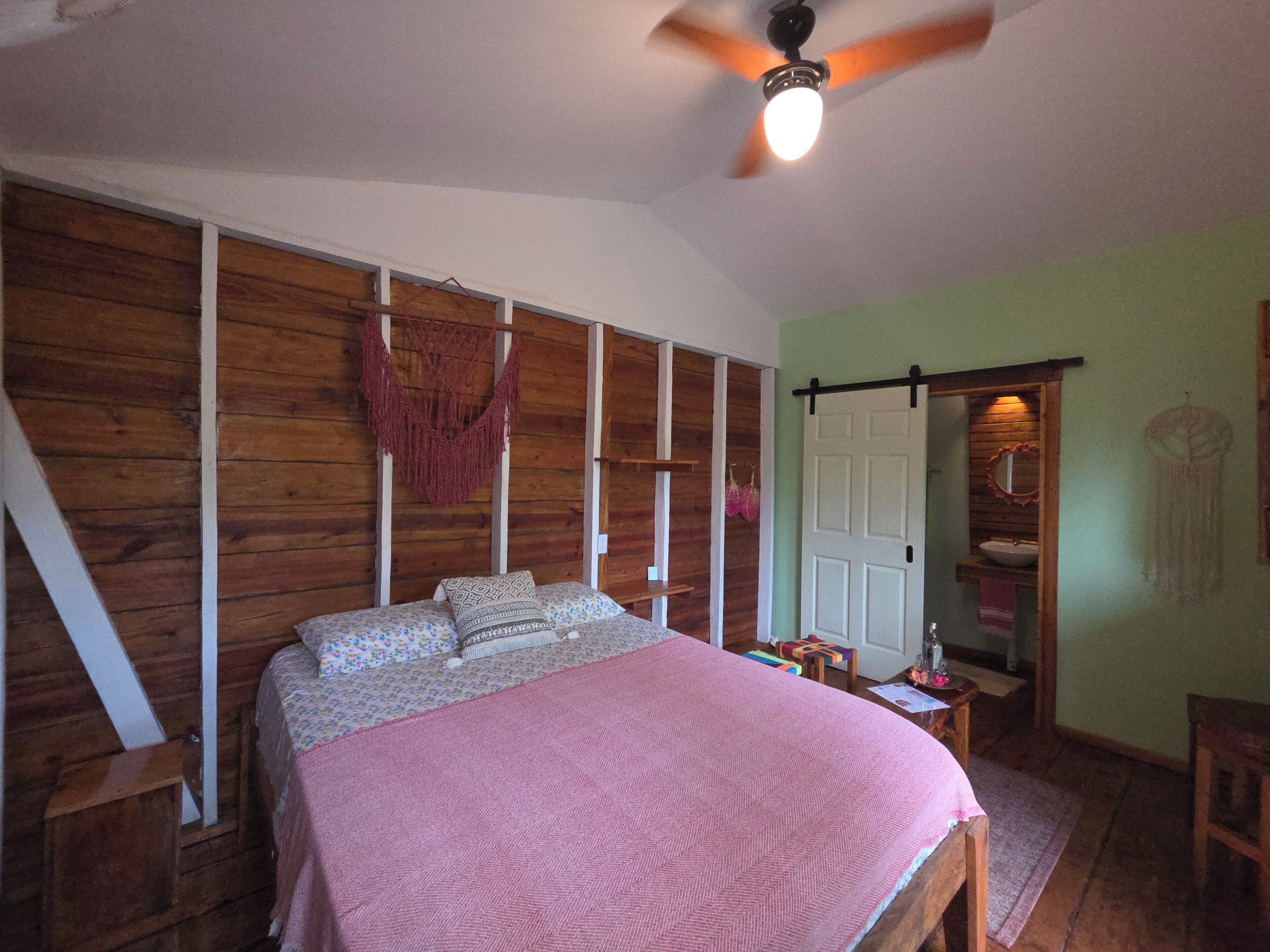 Quarto com teto de madeira, cama com roupa de cama estampada, ventilador de teto e porta que dá para um banheiro.