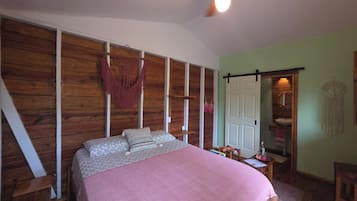 Quarto com teto de madeira, cama com roupa de cama estampada, ventilador de teto e porta que dá para um banheiro.