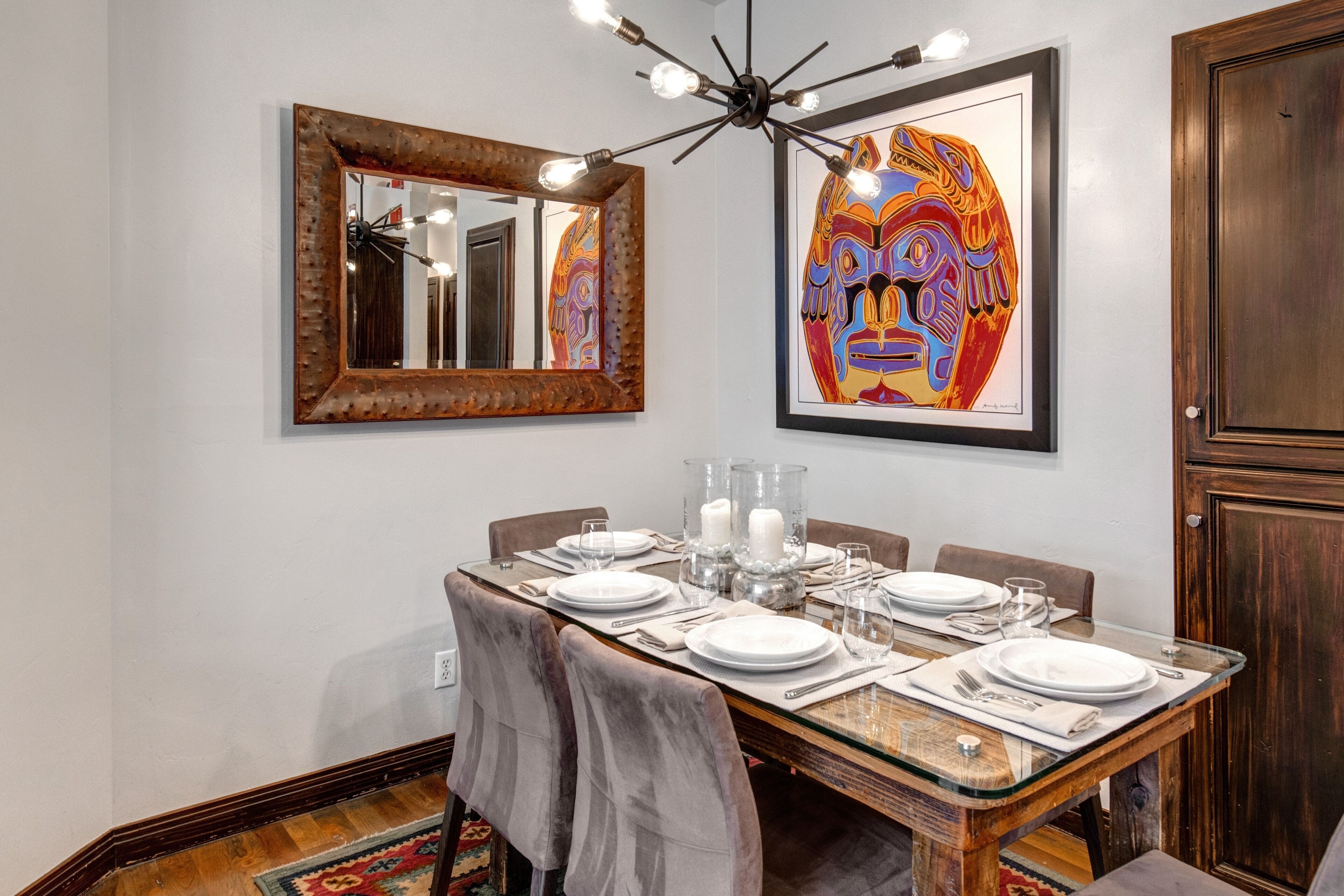 Uma sala de jantar com uma mesa de madeira posta para seis pessoas, um espelho e uma obra de arte colorida na parede.