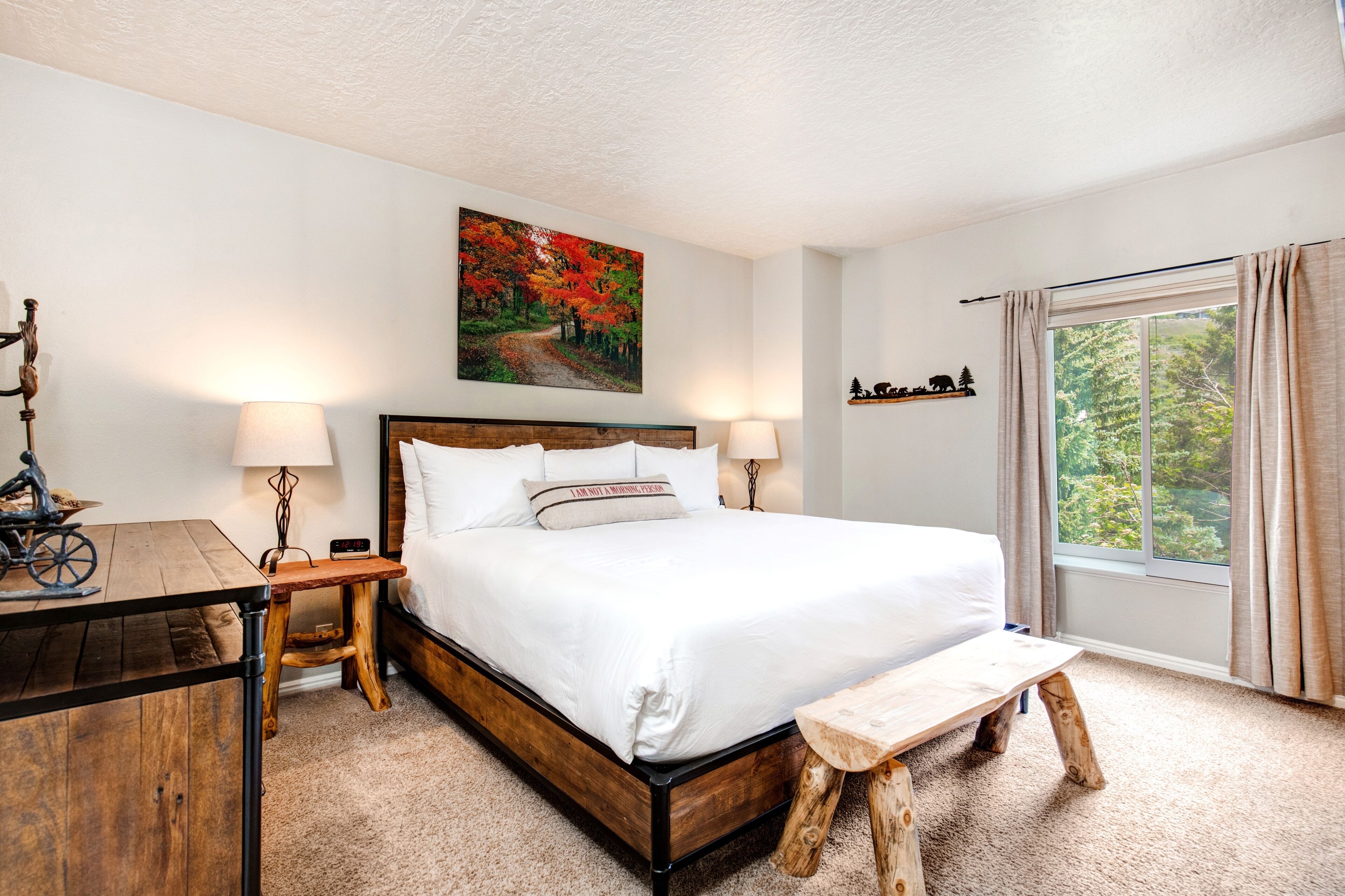 Quarto com estrutura de cama de madeira, roupa de cama branca, mesa de cabeceira com abajur, um banco, pintura de árvores de outono e janela com cortinas.