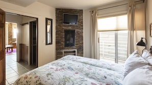 Quarto com cama, televisão montada em parede de pedra, janela com cortinas e porta para outro cômodo.