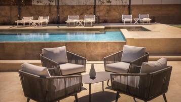 Uma área à beira da piscina com cadeiras de vime e uma pequena mesa, rodeada por espreguiçadeiras e uma piscina.