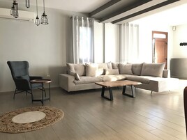 Uma sala de estar com um sofá de canto, uma mesinha redonda, uma cadeira e uma mesa de centro.