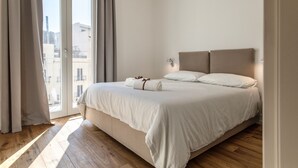 Quarto com cama grande, janela com cortinas e vista para prédios.