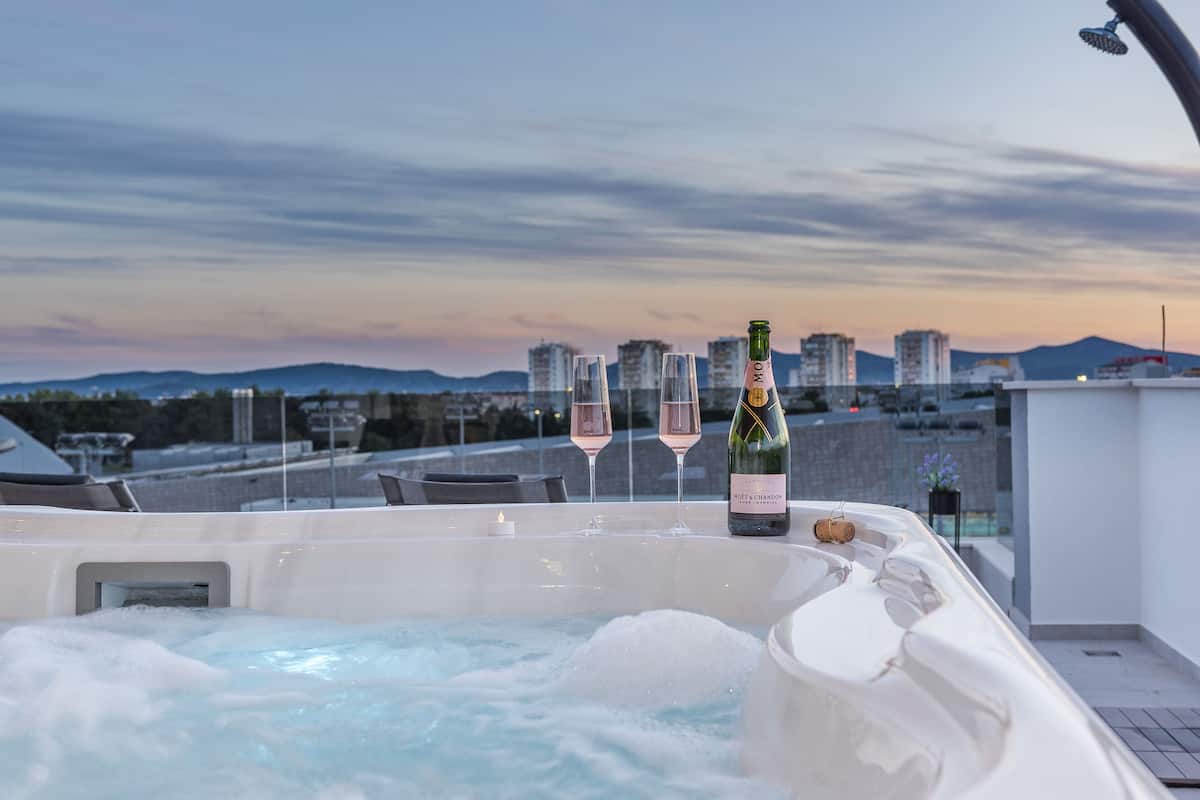 Uma banheira de hidromassagem com taças de champanhe e uma garrafa, com vista para um horizonte urbano ao pôr do sol.