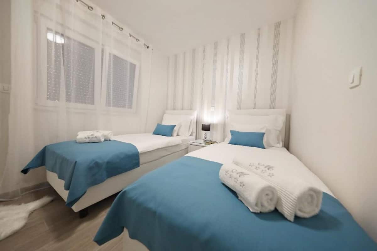 Quarto de hotel com duas camas, cada uma com colcha azul e almofadas brancas. Há uma janela com cortinas, uma mesa de cabeceira com abajur e um tapete branco no chão.