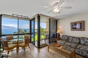 Quarto com porta de vidro que dá para uma varanda com vista para o oceano, ventilador de teto, sofá e mesa com cadeiras.