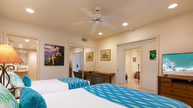 Quarto de hotel com duas camas, um ventilador de teto, uma escrivaninha com cadeira, televisão e obras de arte nas paredes.