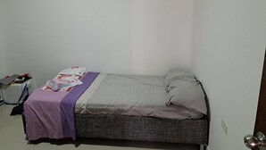 Quarto pequeno com uma cama de solteiro, colchão cinzento e manta roxa. Há uma pequena mesa com uma cadeira à esquerda, e uma porta com puxador prateado à direita.