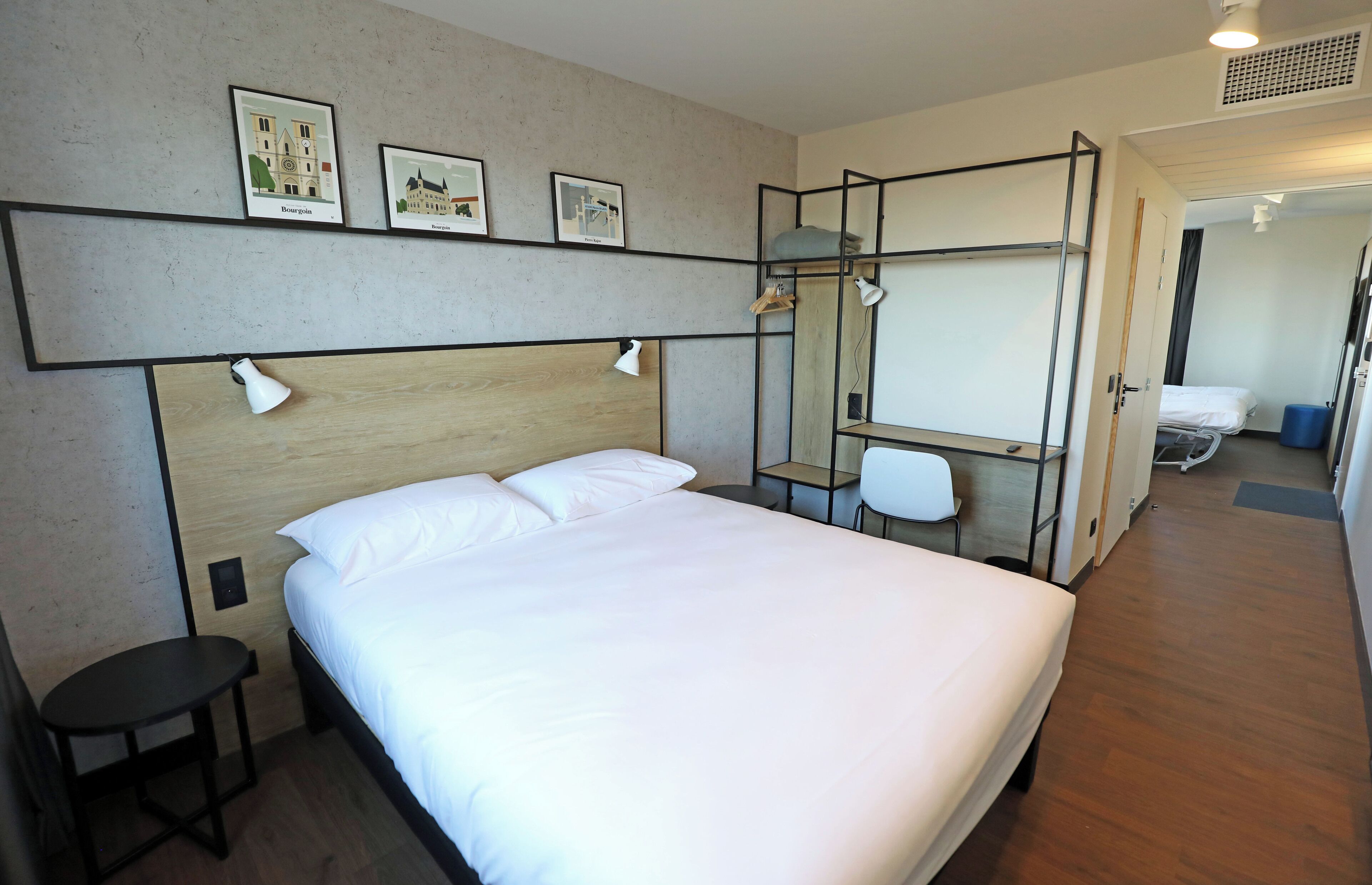 Quarto de hotel com uma cama grande, uma escrivaninha e uma cadeira. Há três quadros na parede acima da cama.