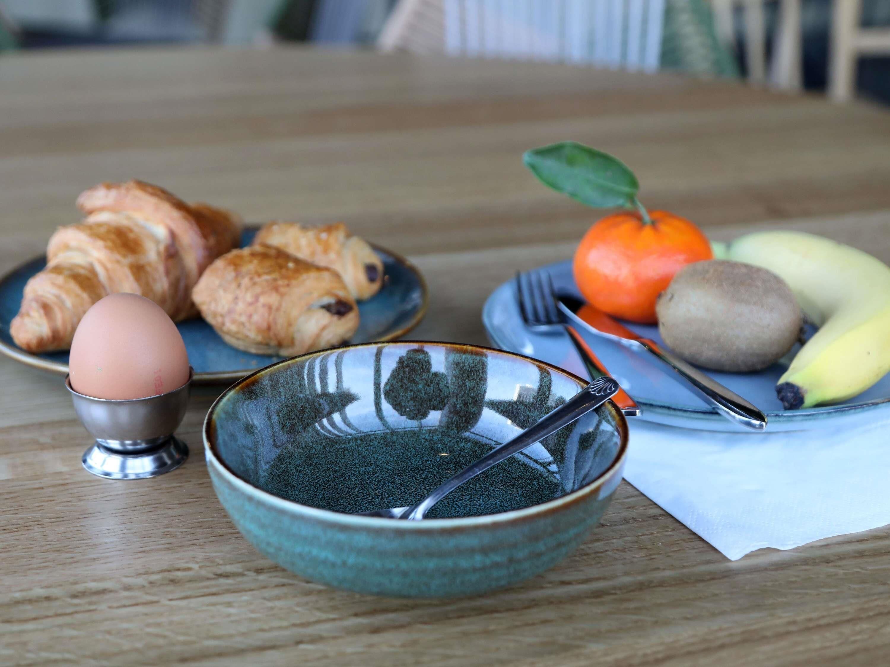 Disposição de café da manhã com croissants, um ovo, uma tigela de chá verde, uma laranja, um kiwi e uma banana sobre uma mesa de madeira.