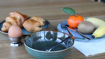 Disposição de café da manhã com croissants, um ovo, uma tigela de chá verde, uma laranja, um kiwi e uma banana sobre uma mesa de madeira.