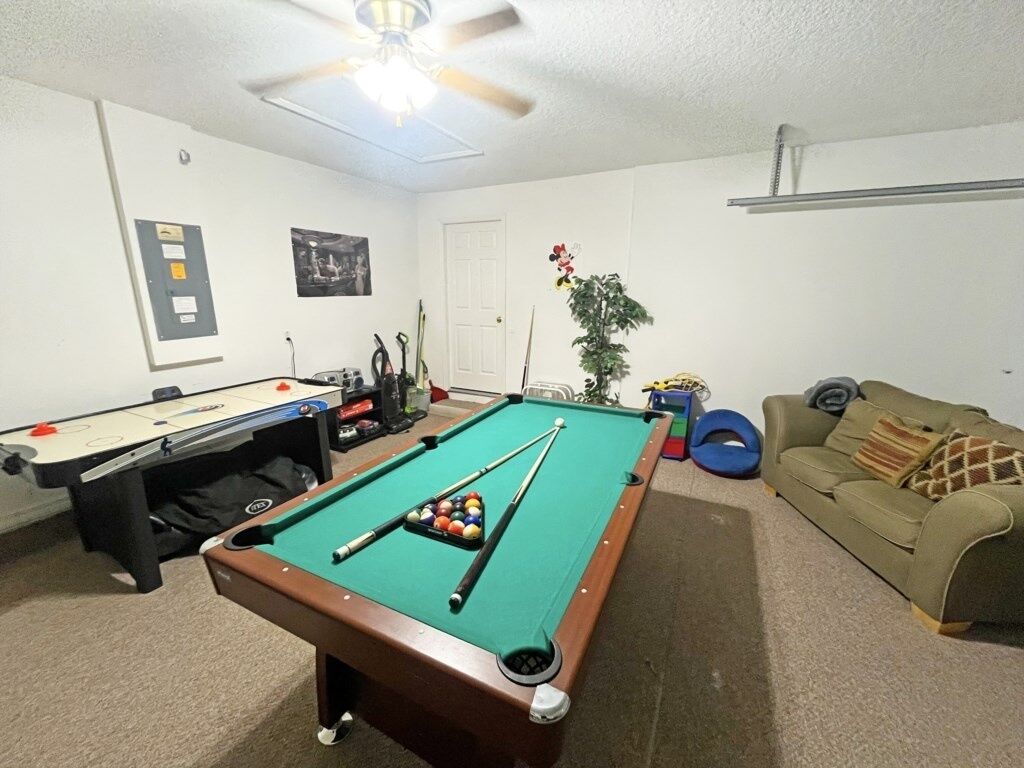 Uma mesa de bilhar com bolas, um sofá e um ventilador de teto em um quarto com paredes brancas.