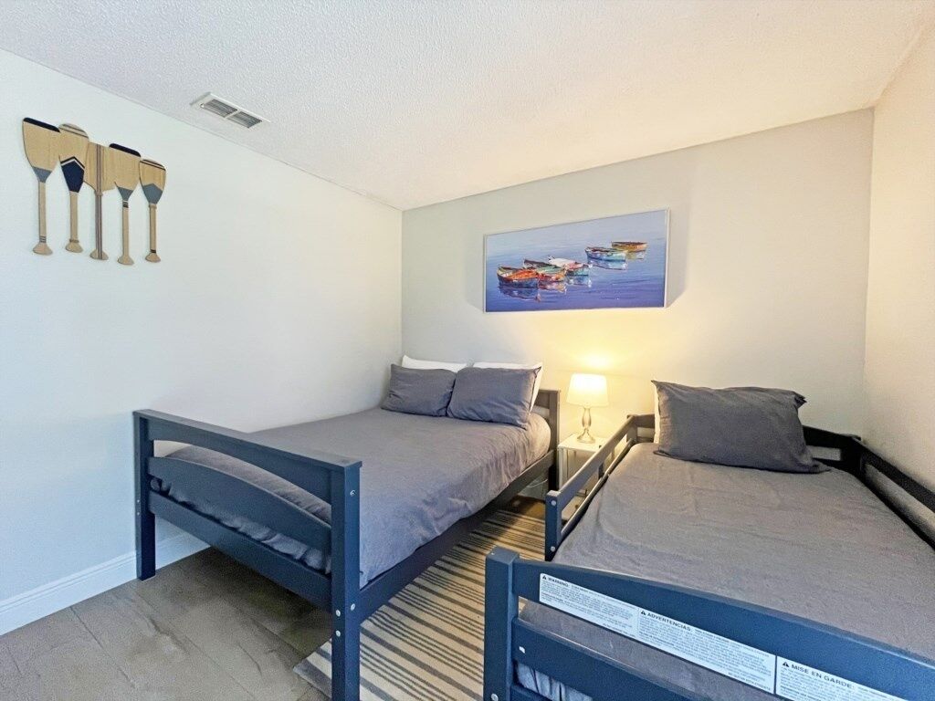 Quarto com duas camas de solteiro, um quadro de barcos, uma luminária na parede e um respiradouro no teto.