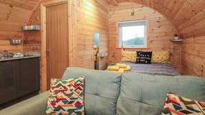 Uma cabana acolhedora com interior de madeira, uma cama com duas almofadas, uma janela e uma cozinha compacta.