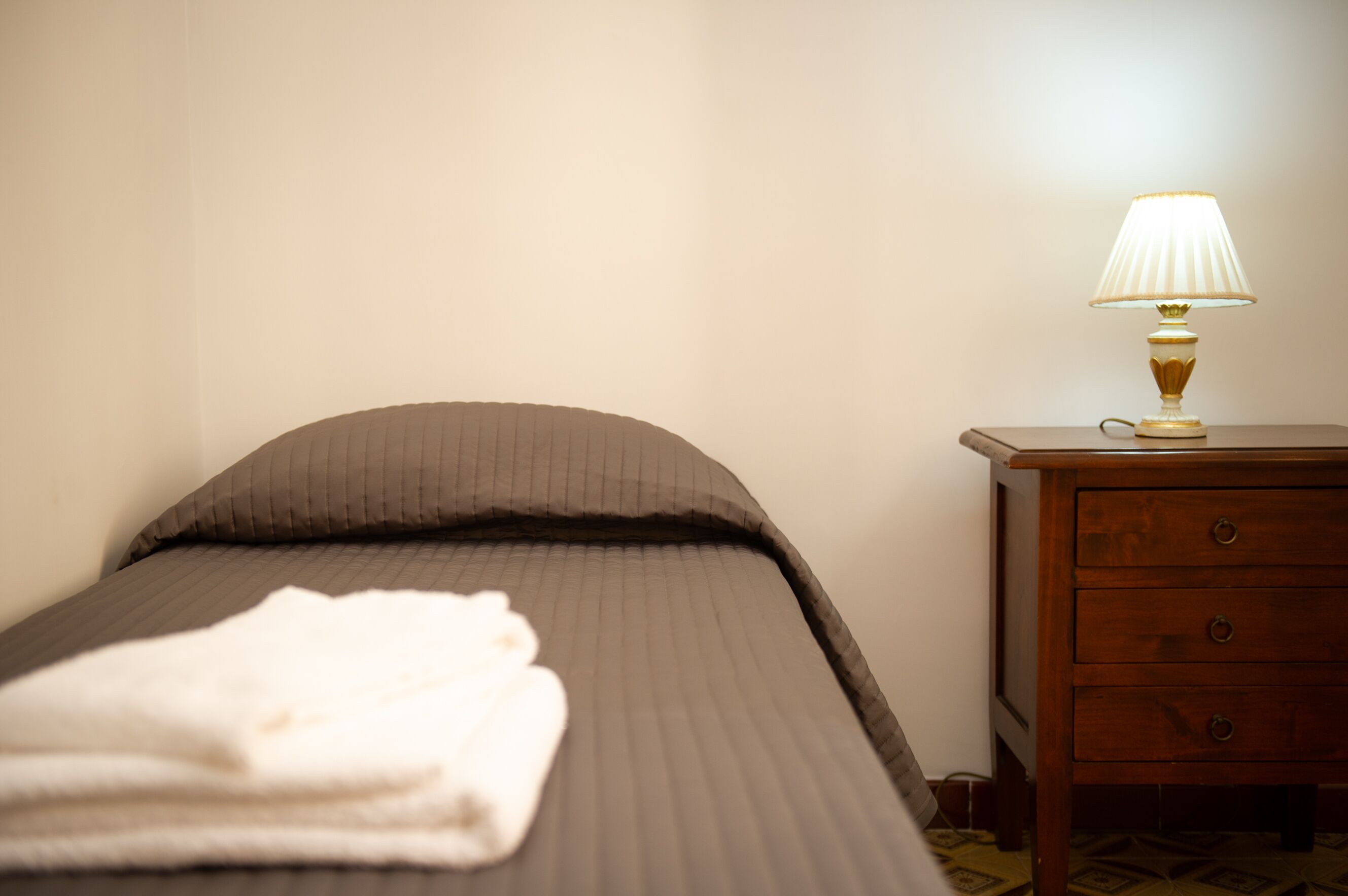 Uma cama de solteiro com cabeceira marrom e manta branca, um criado-mudo de madeira com abajur, e uma cômoda de madeira com gavetas.