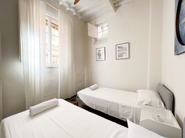 Quarto com duas camas de solteiro, uma janela com cortinas leves e dois quadros na parede.