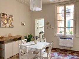 Uma sala de jantar com uma mesa e cadeiras brancas, uma luminária pendente branca, uma janela com vista para o exterior e um radiador na parede.