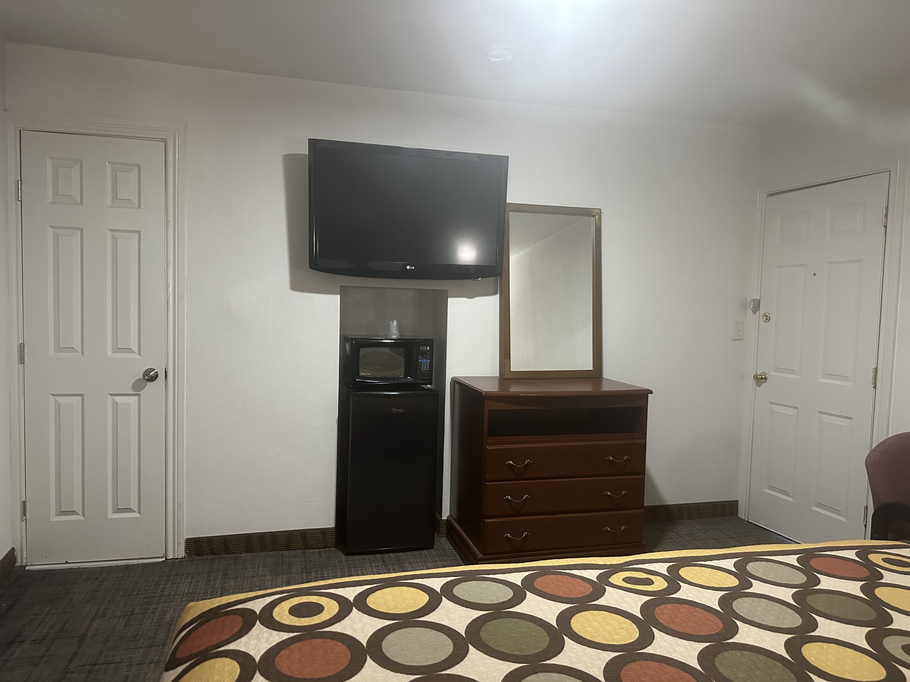 Quarto de hotel com televisão de tela plana acima de uma geladeira, penteadeira com gavetas e colcha estampada.
