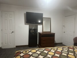 Quarto de hotel com televisão de tela plana acima de uma geladeira, penteadeira com gavetas e colcha estampada.