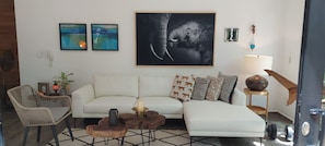 Uma sala de estar moderna com um sofá branco, uma poltrona de vime e uma mesa de centro. Há duas obras de arte emolduradas acima do sofá e uma grande pintura de elefante na parede.